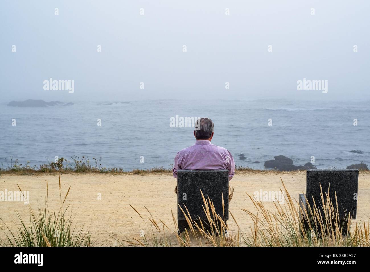 Lonely man seated on a stone bench looking at the Ocean in a foggy day ...