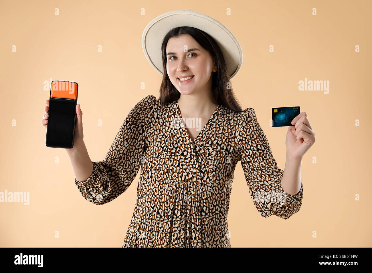 Happy young woman with payment terminal and debit card on beige ...