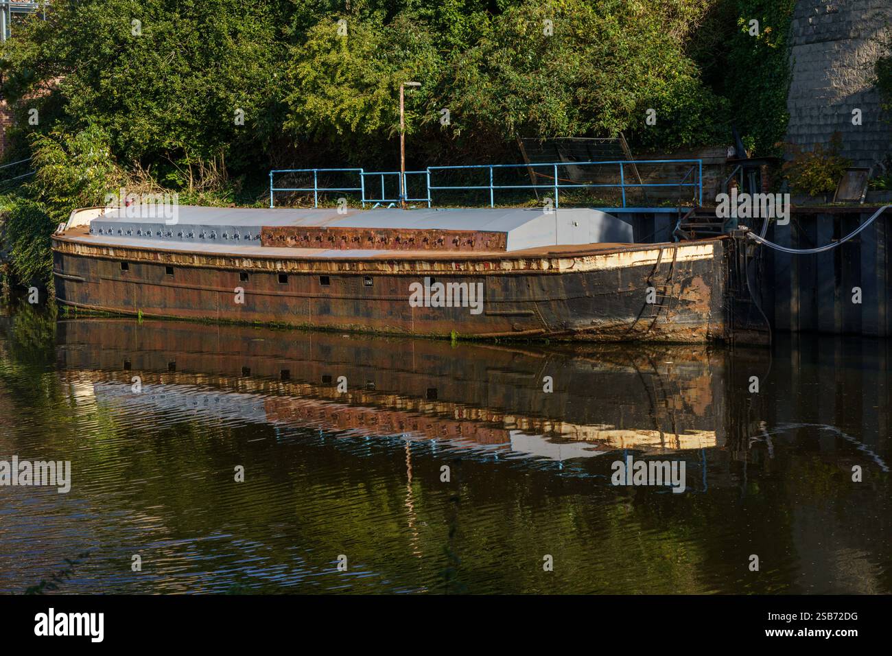 A weathered barge sits still on a waterway, its reflection mirrored in ...