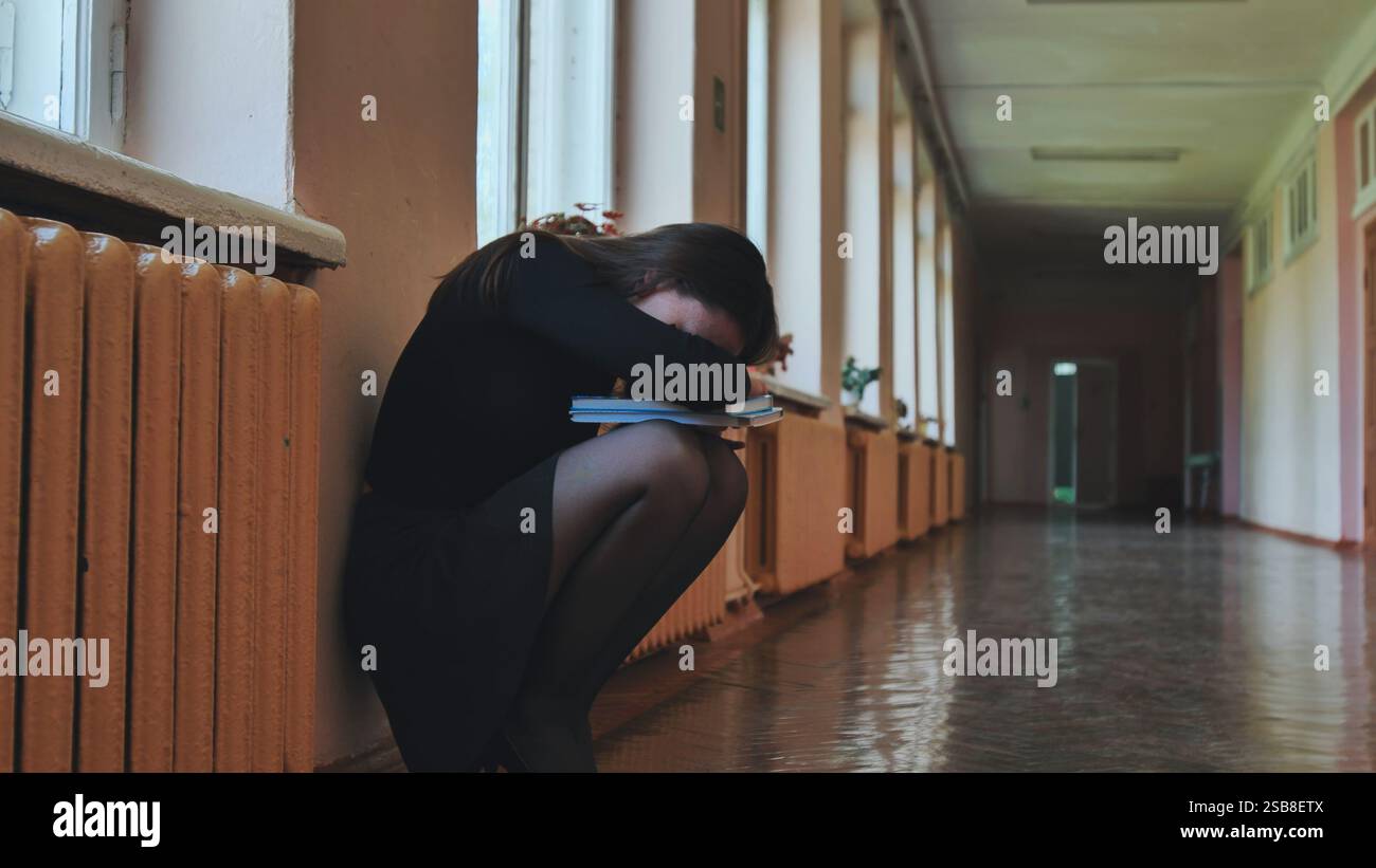 Teenage student crouching on the floor in school hallway, hugging a ...