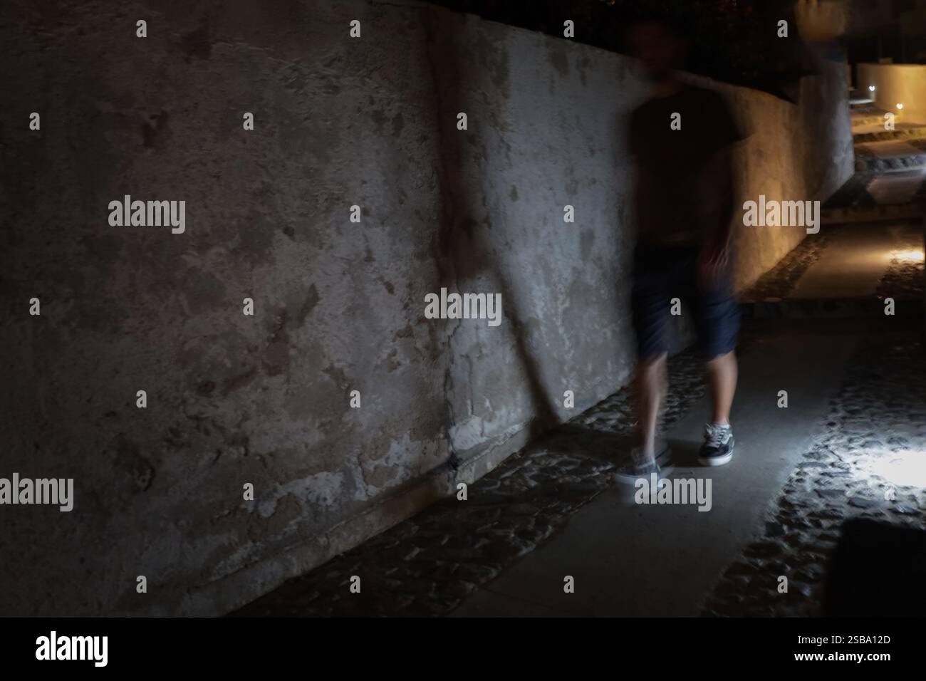 A blurred figure walks on a dimly lit path next to a textured concrete ...