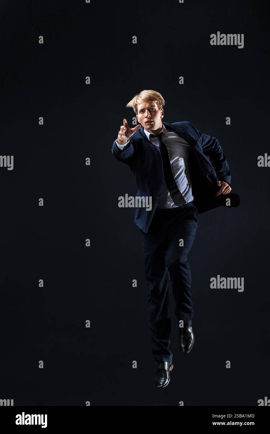In a dark studio, a man in a sharp navy suit leaps mid-air, reaching ...