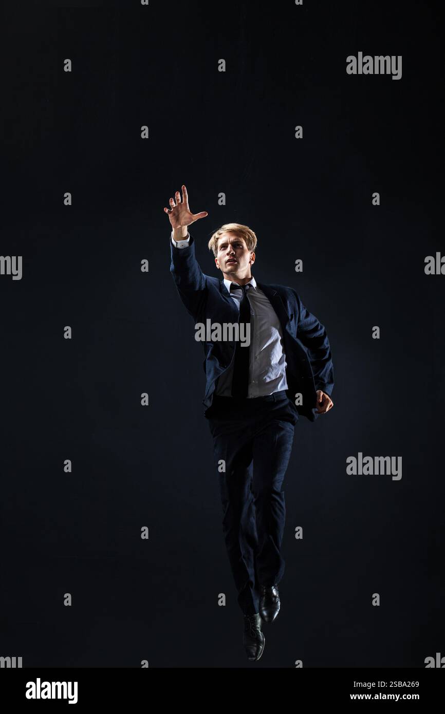 In a dark studio, a man in a sharp navy suit leaps mid-air, reaching ...