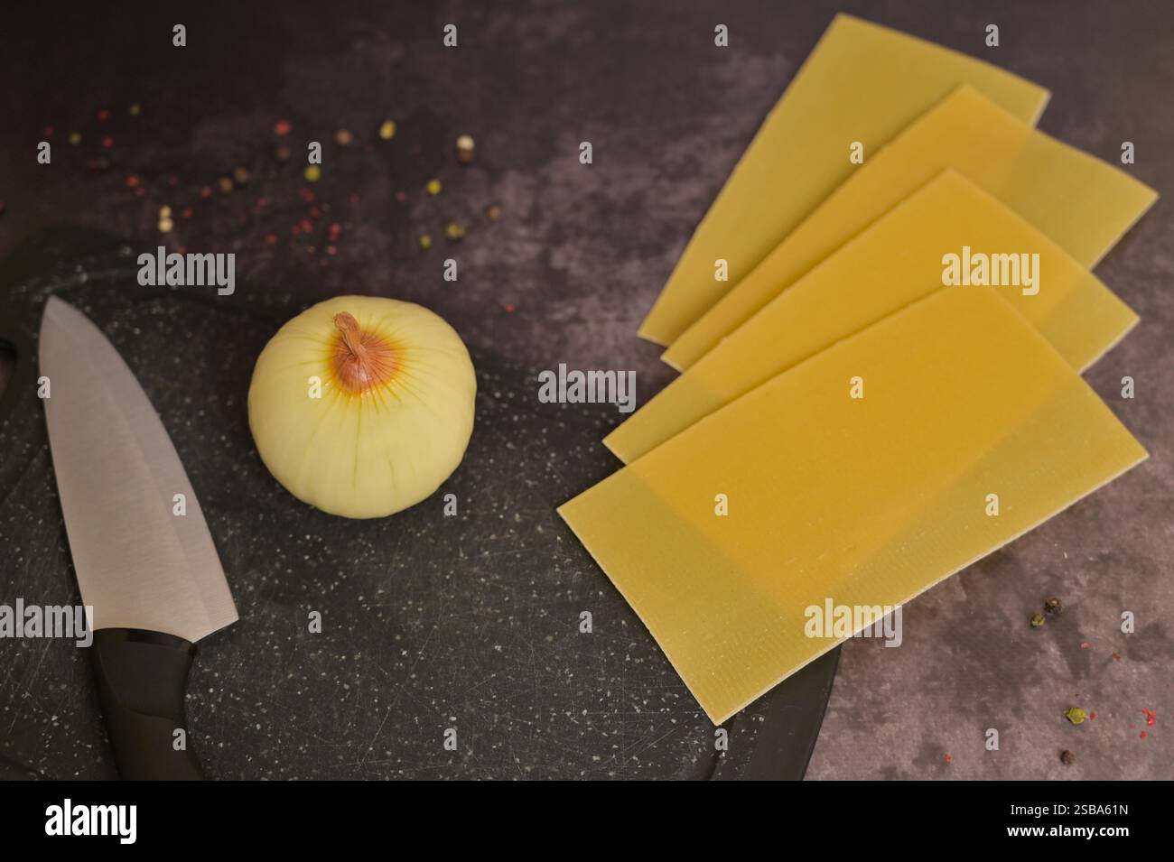Onion, knife and lasagne pasta sheets on a black kitchen cutting board ...