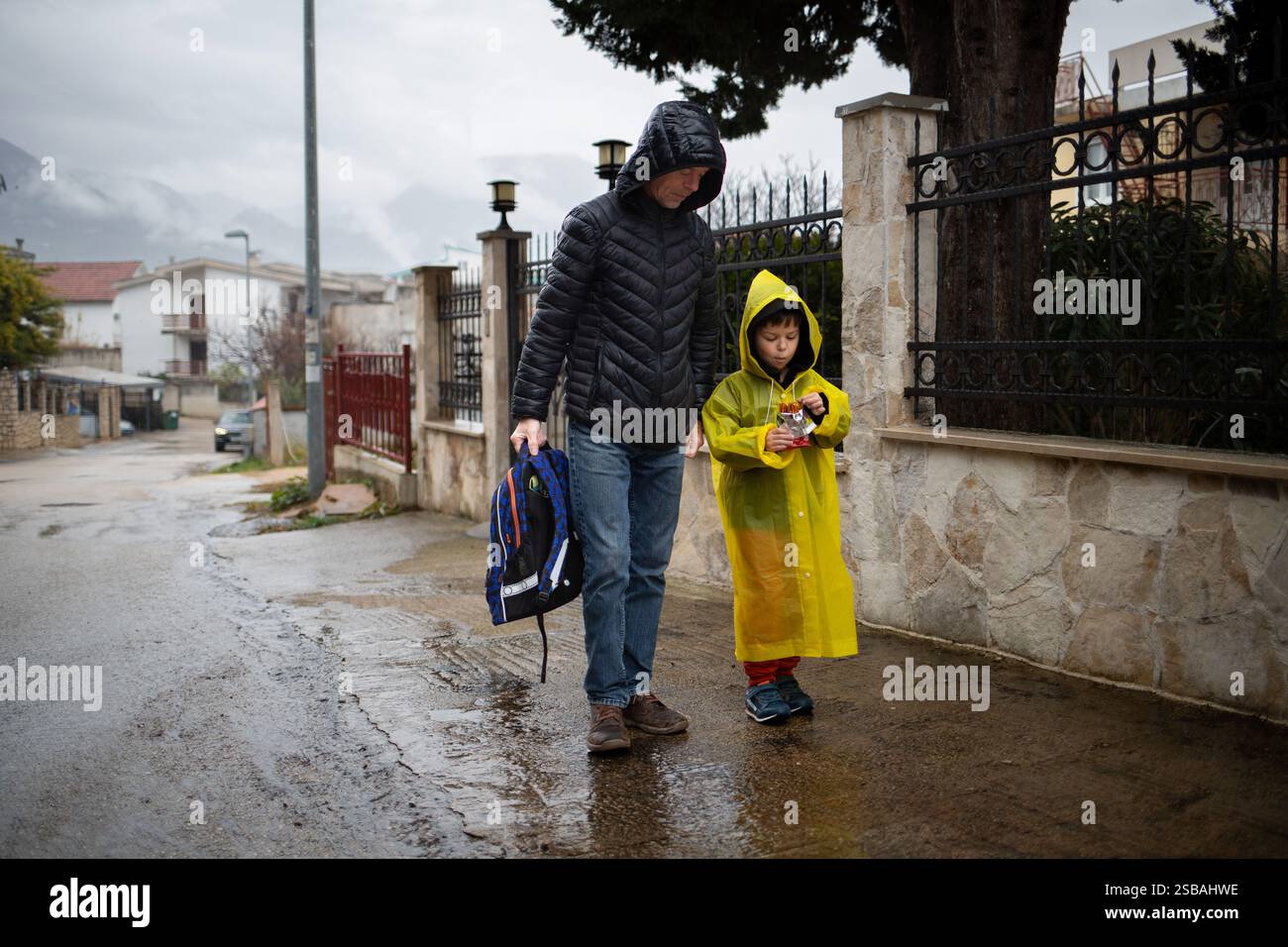 Father and son walk in rain after school. Child in yellow raincoat eats ...