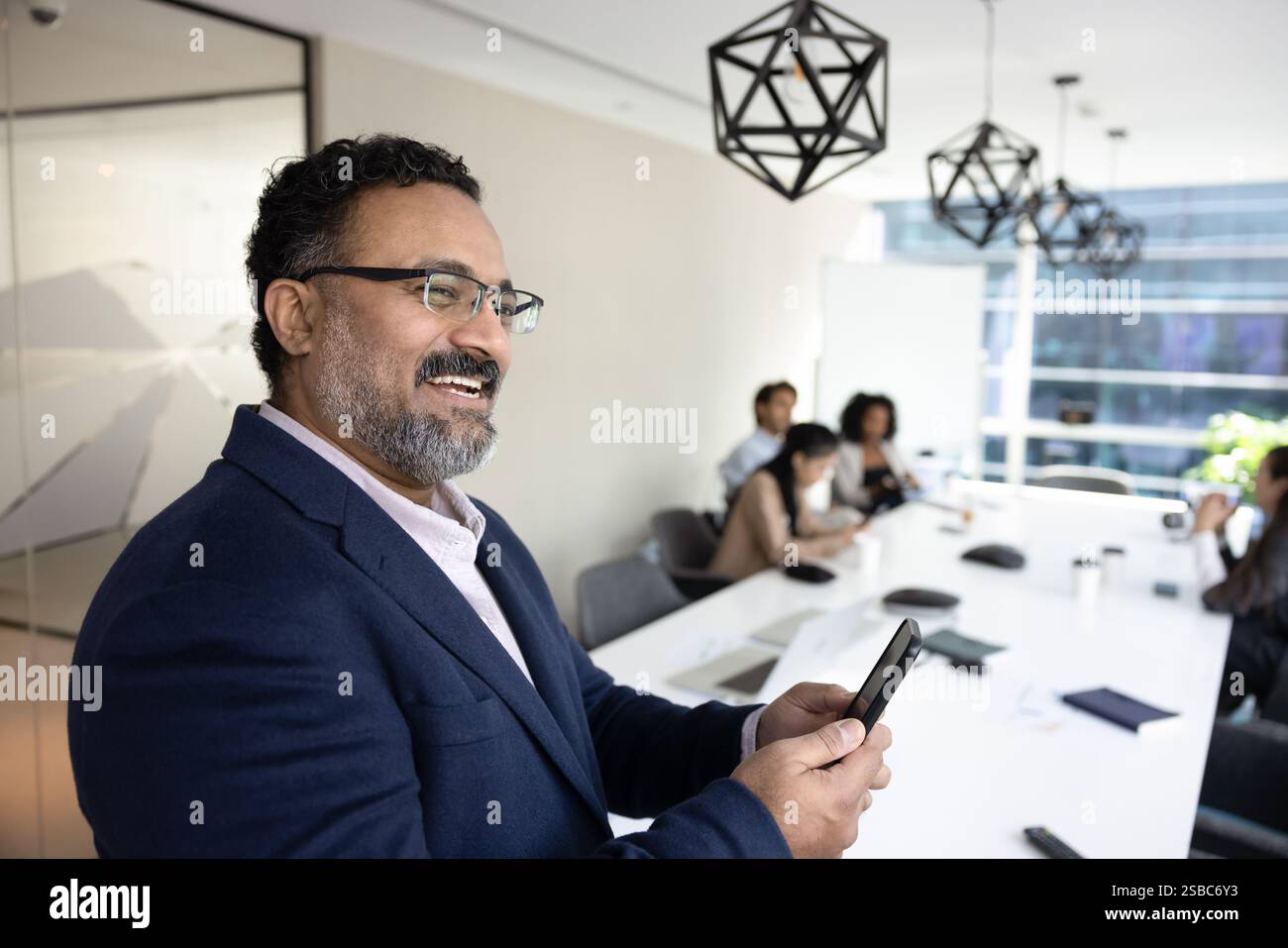 Cheerful mature Indian CEO with smartphone standing in meeting room ...