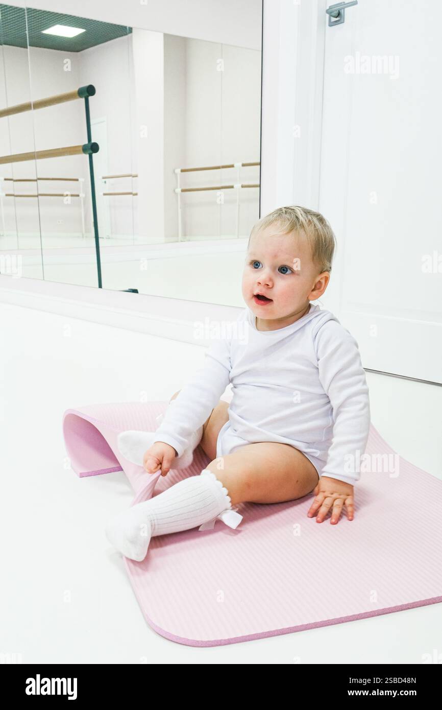 Portrait of a cute baby ballerina sitting near the ballet barre putting ...