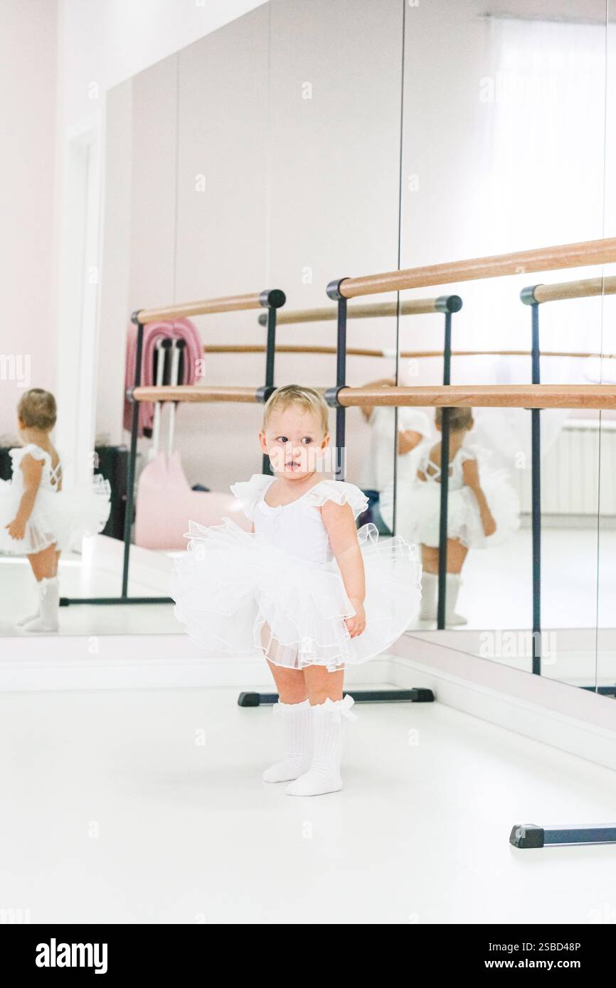 Portrait of a cute baby ballerina sitting near the ballet barre putting ...