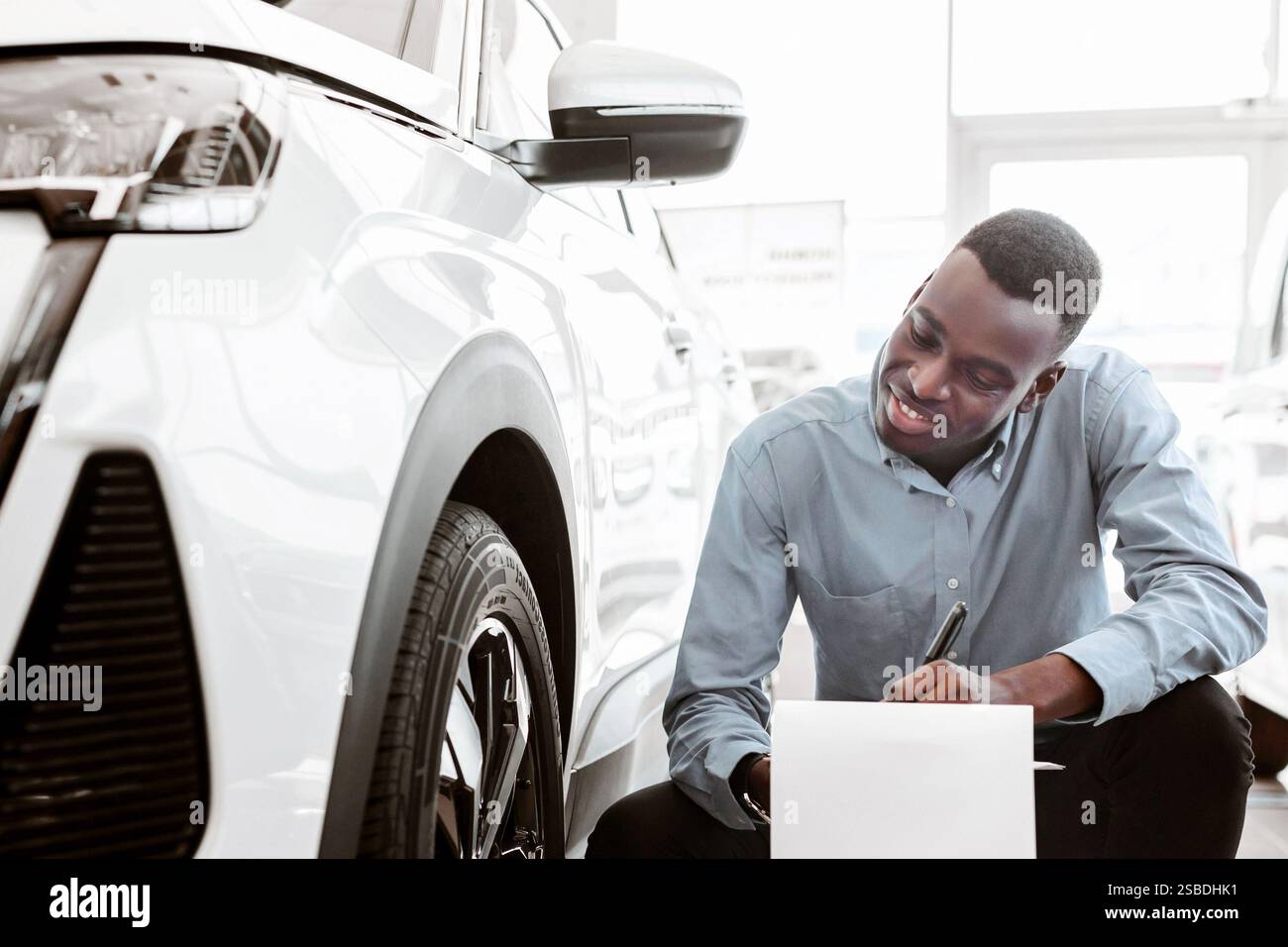 Smiling black car salesman sitting near new auto, writing in clipboard ...