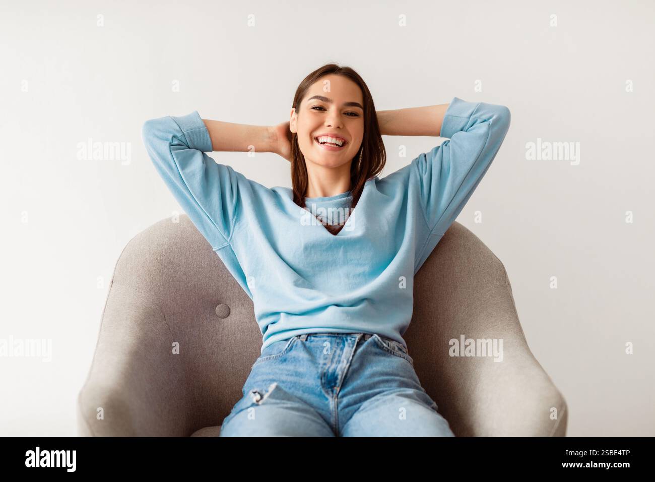 Young woman sitting in armchair, holding hands behind head, resting and ...