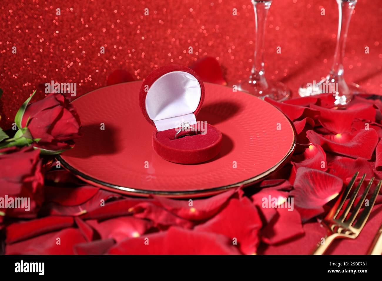 Engagement ring in box, tableware, rose flowers and petals on red glitter background, closeup ...