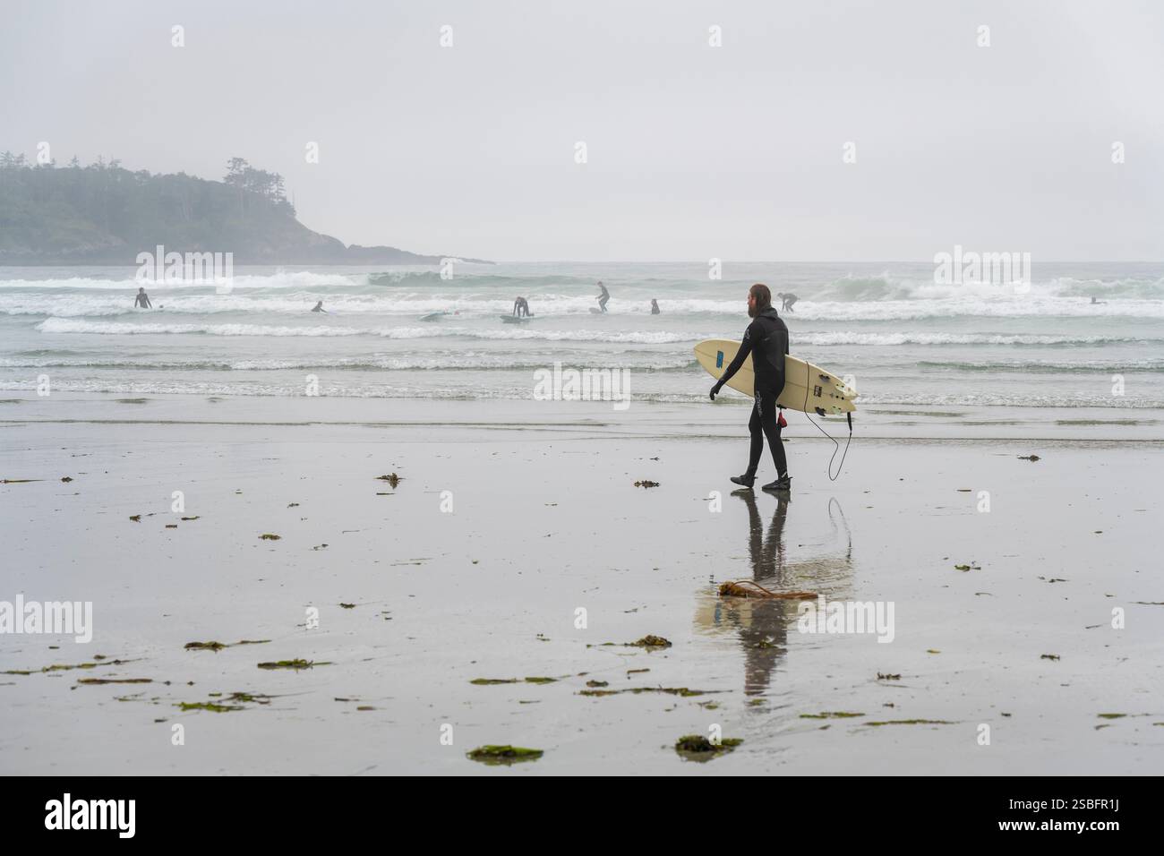 Young active man with surfboard walking on Cox Bay Beach and surfers in ...
