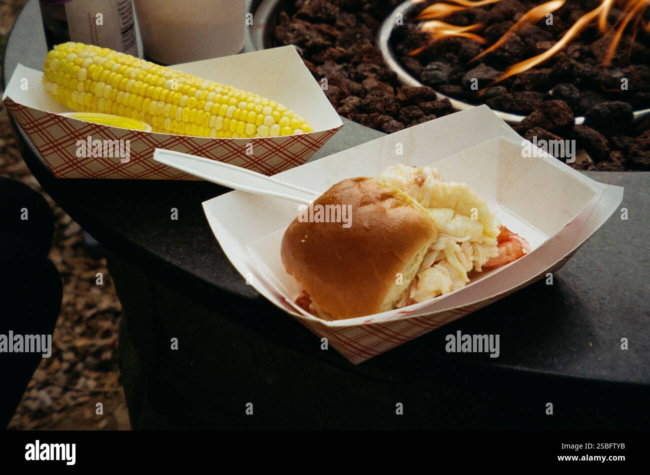 Lobster roll and corn on the cob served in paper trays by a fire pit ...