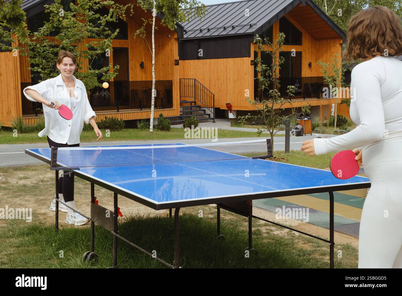 Two women play an outdoor game of table tennis, surrounded by wooden ...
