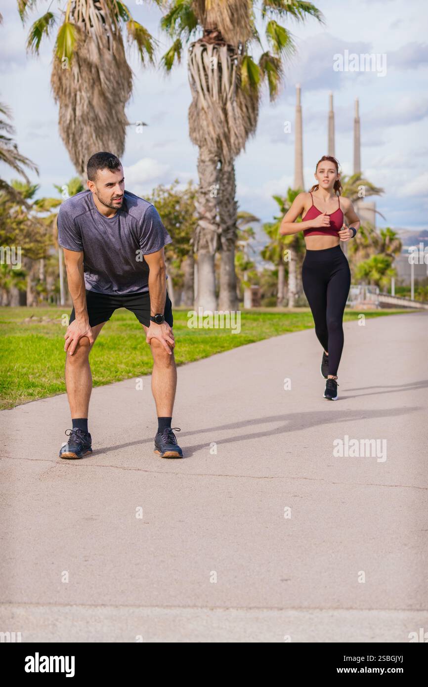 Tired man resting after jogging while woman continues running in a park ...