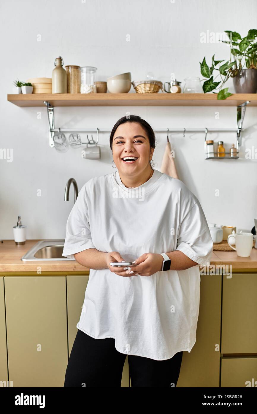 A young plus sized woman smiles in a lovely kitchen, holding her phone ...