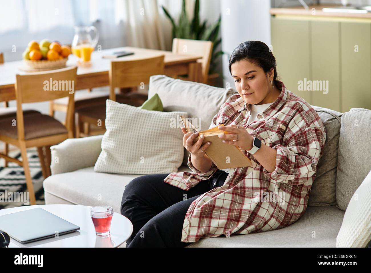 A young plus sized woman sits comfortably on a couch reading a book ...