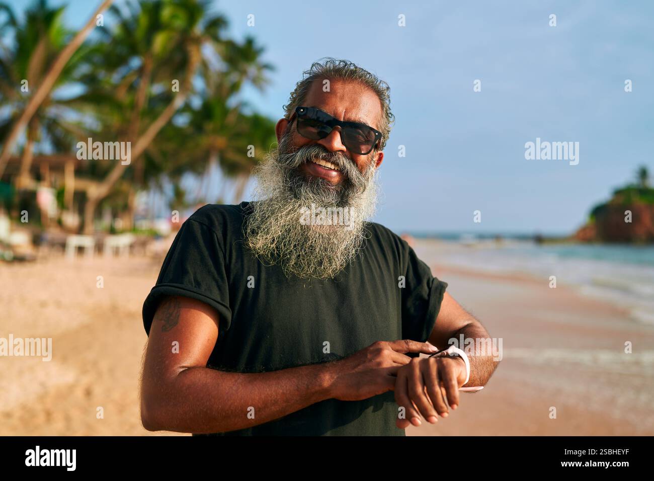 Stylish gray-bearded senior with sunglasses uses smart watch on tropical beach. Modern tech ...
