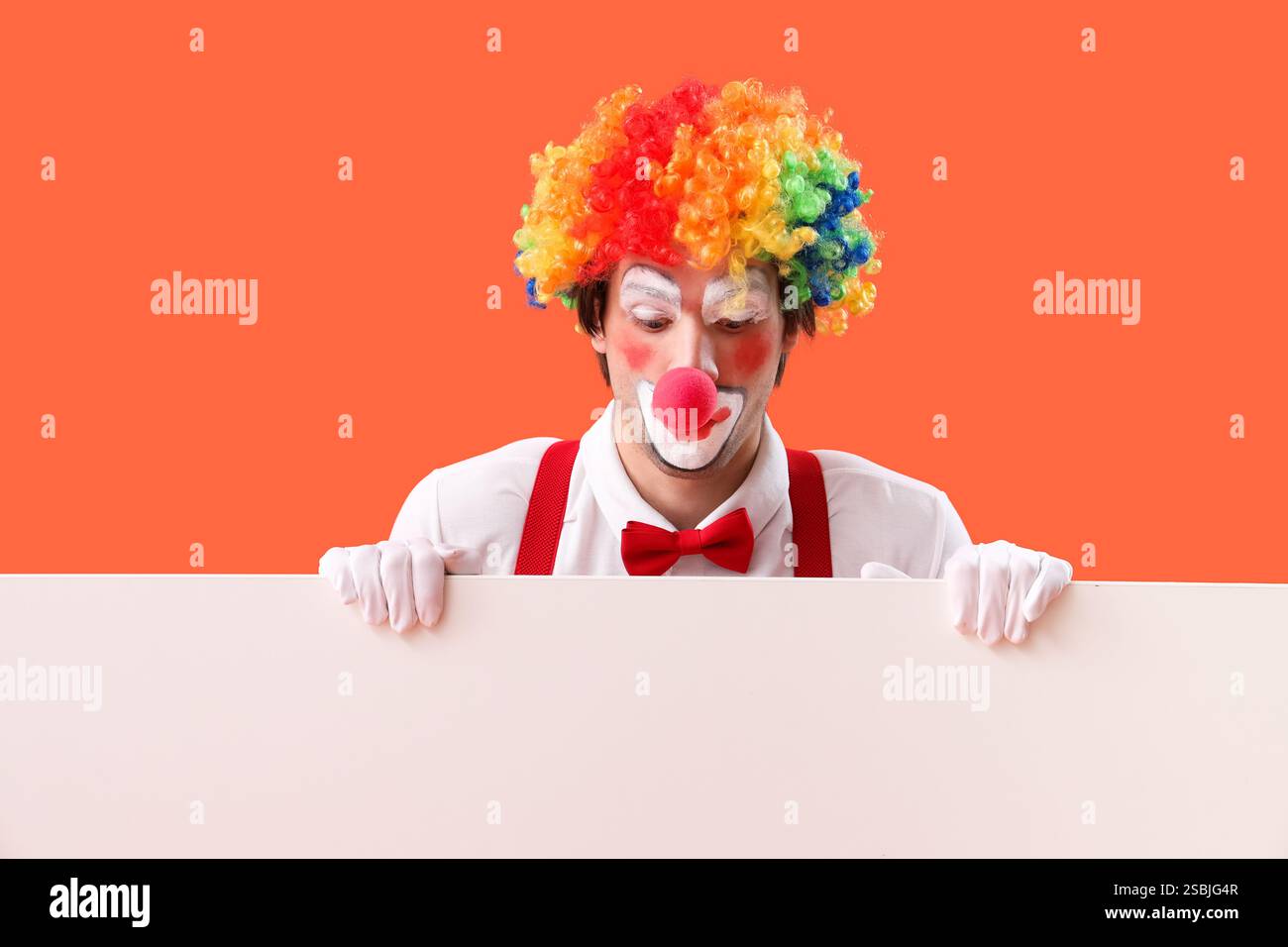 Portrait of surprised clown with blank poster on orange background ...