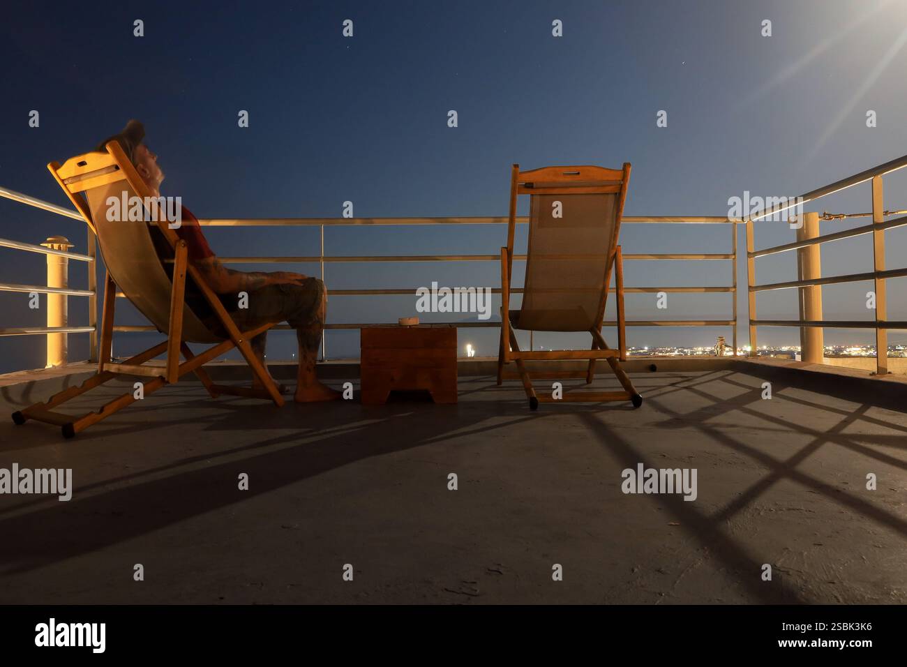 A person sits in a wooden chair on a rooftop balcony at night, basking ...