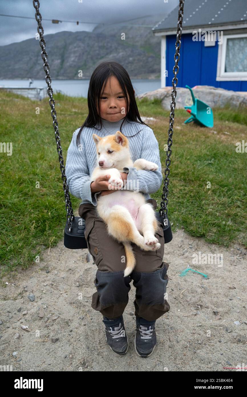 A Little Inuit girl sits on a swing, cradling a playful puppy in ...
