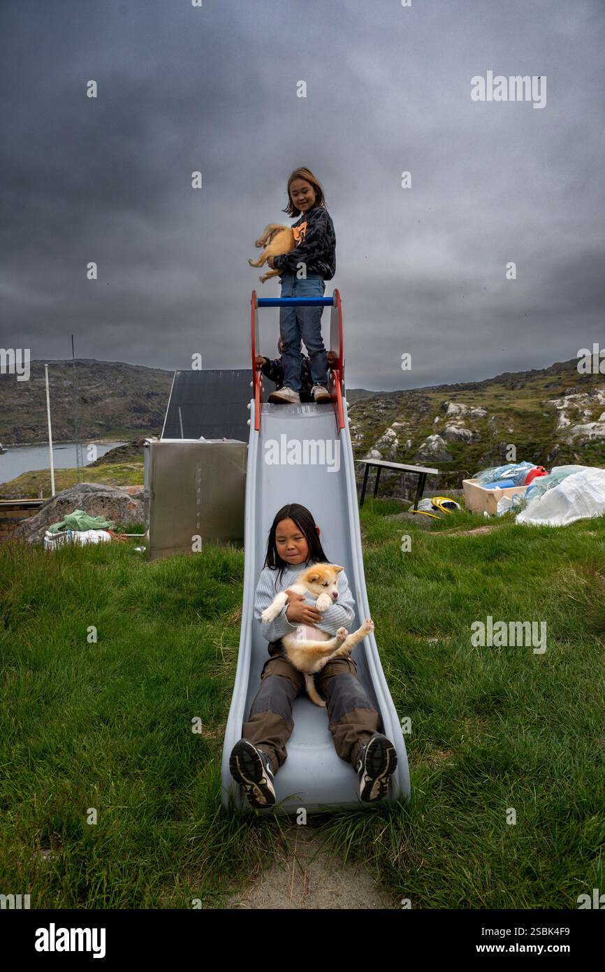 Two Inuit children engage in play on a slide while holding dogs in the ...