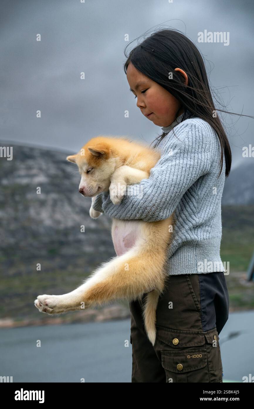 Little Inuit girl gently cradles a small puppy by the scenic shores of ...