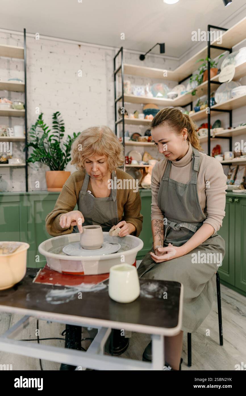 Mentor guiding elderly student in sculpting pottery on electric spinning wheel Two generations ...