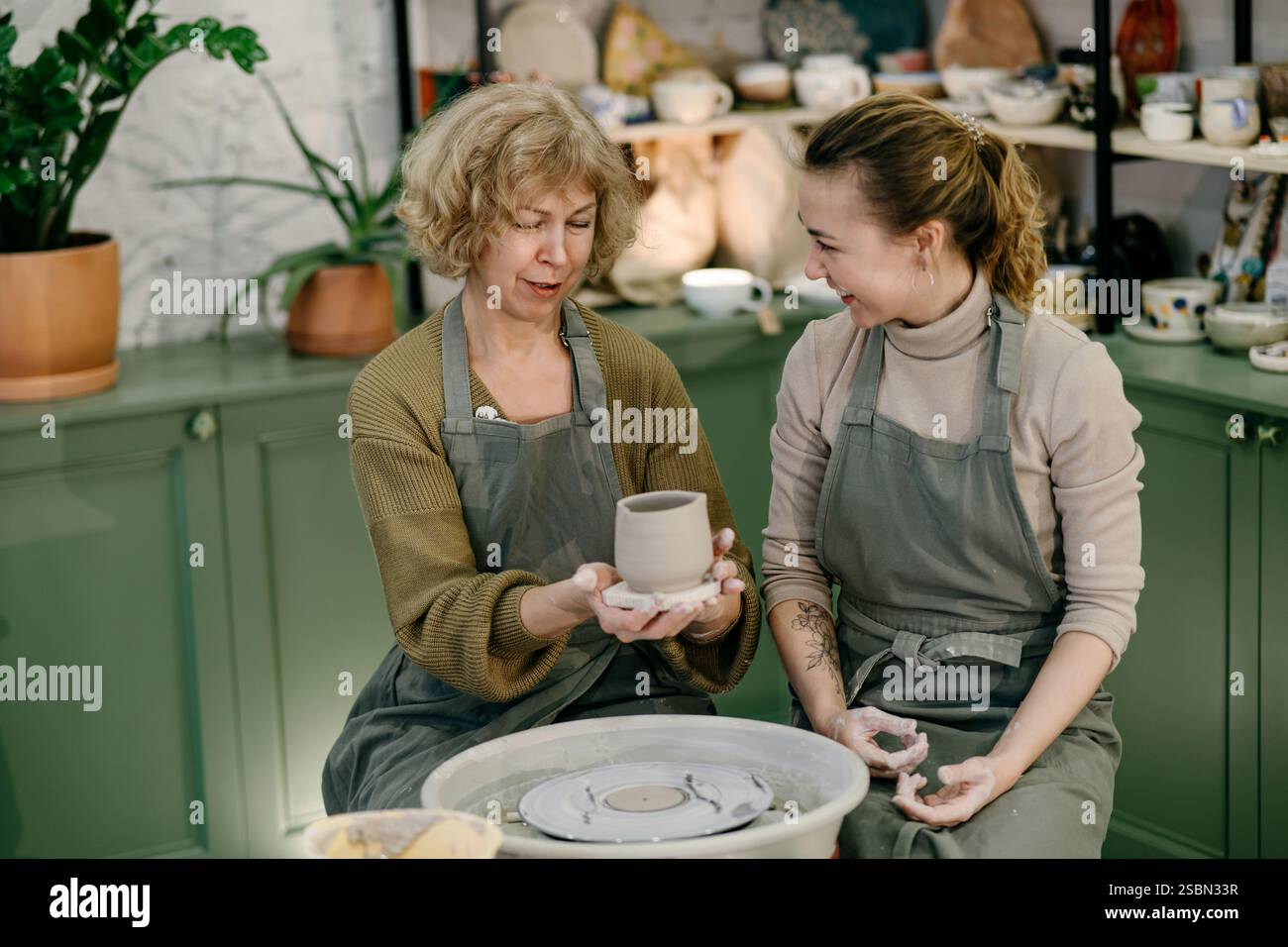 Elderly woman learning pottery on spinning wheel with young instructor ...