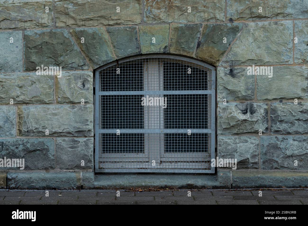 A gray stone wall features a barred window set within an arched opening ...