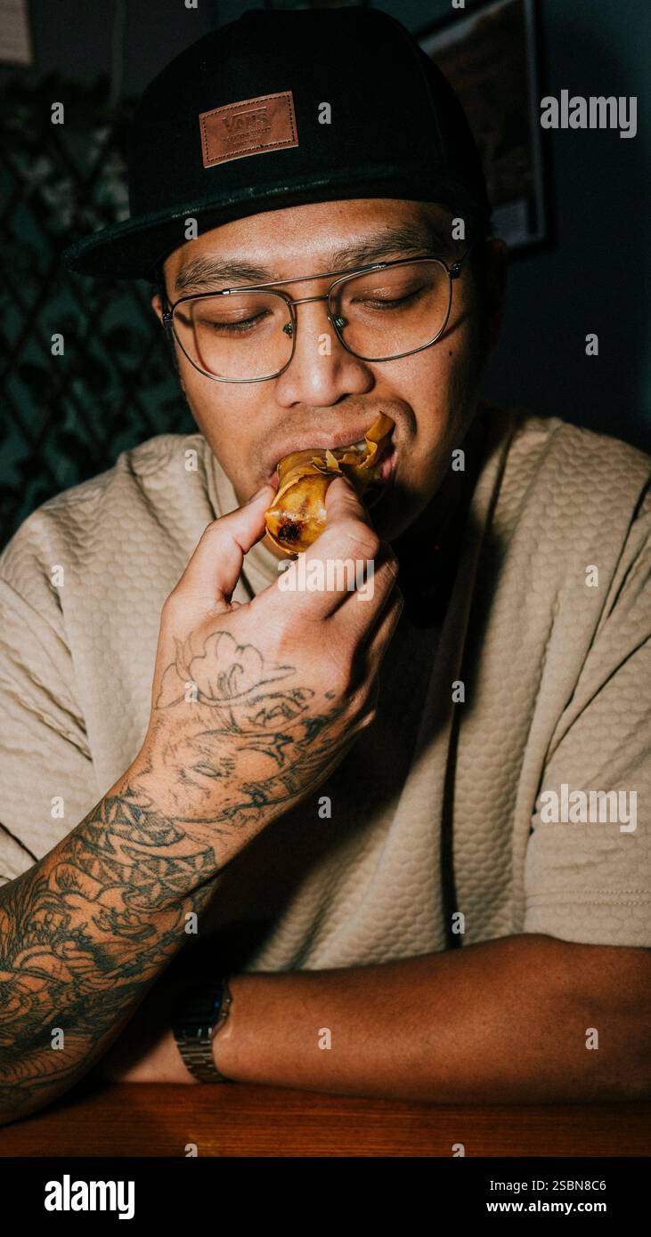A man with tattoos on his arms is eating a spring roll. He is wearing a ...