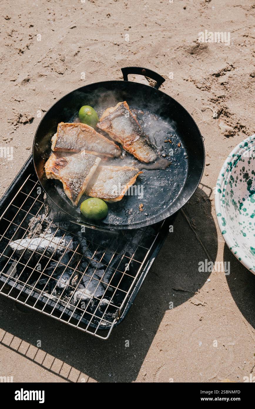 A pan of fish is cooking on a grill on a beach. The fish is accompanied ...