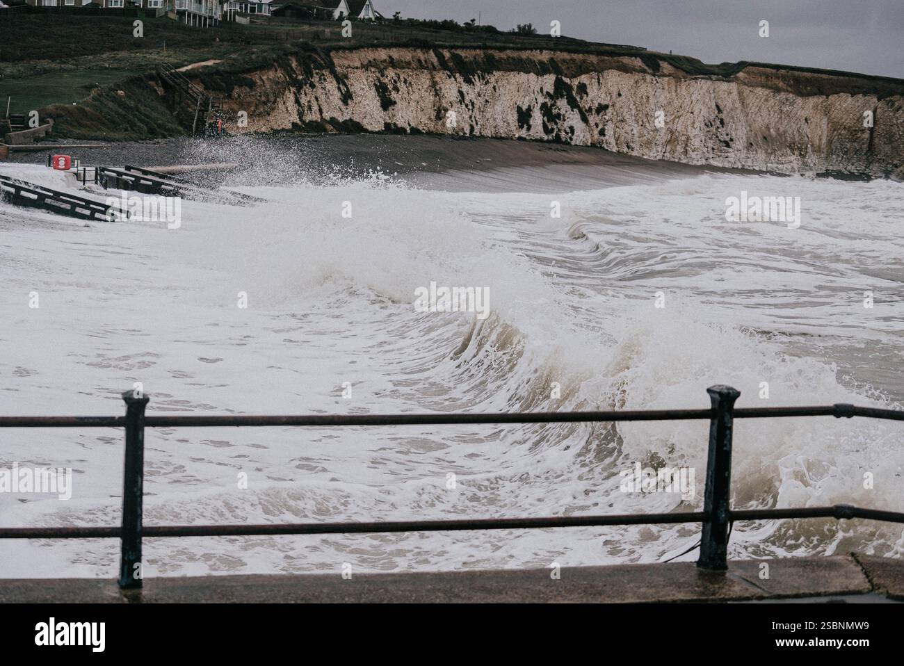 A large wave is crashing into the shore, and a fence is visible on the ...