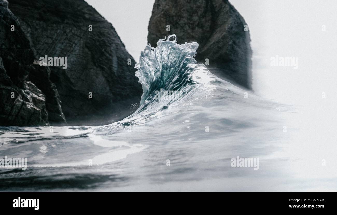 A wave is crashing into a rocky shore. The water is calm and clear, and ...