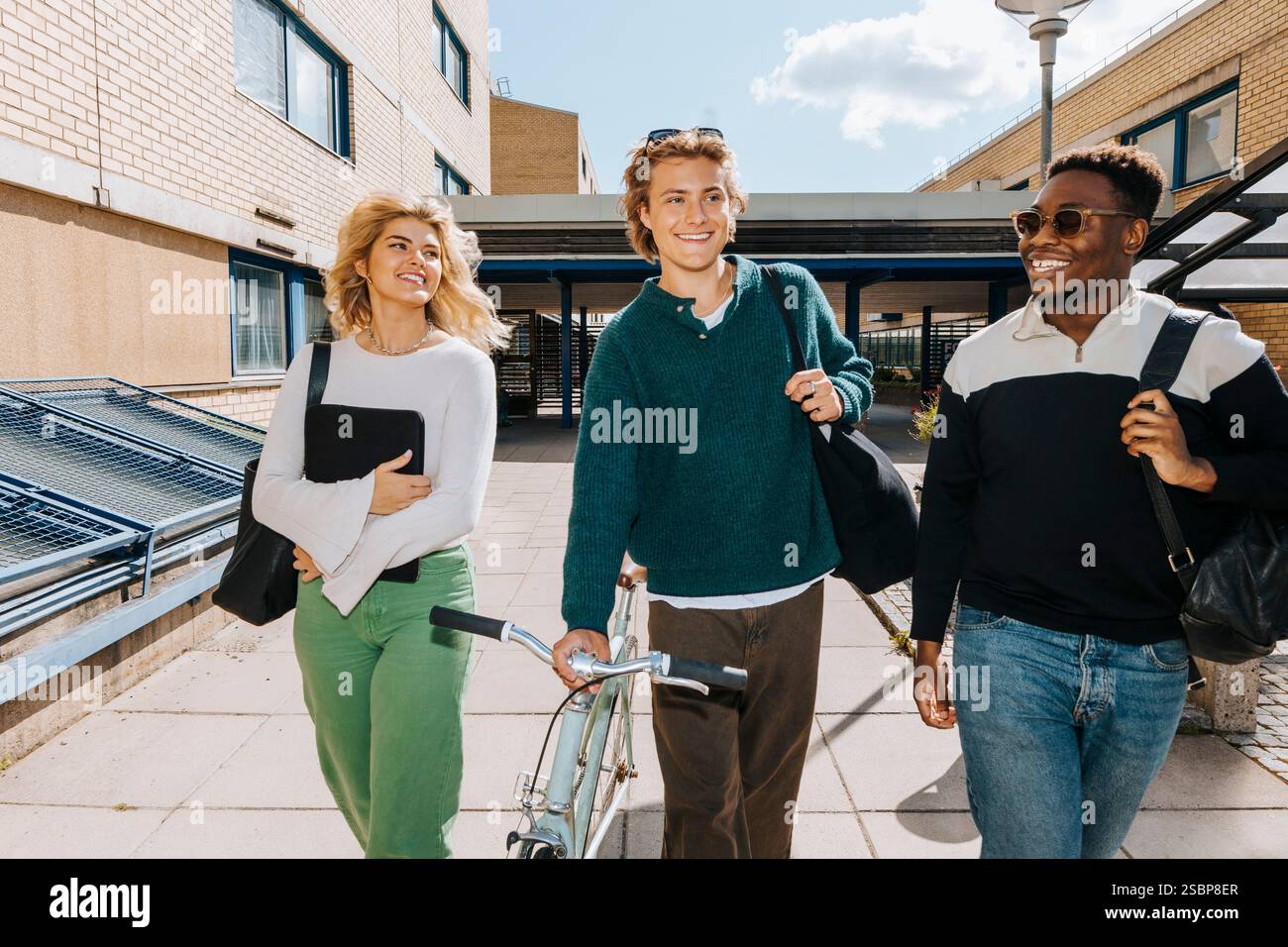 Smiling man wheeling bicycle while walking with friends at college ...