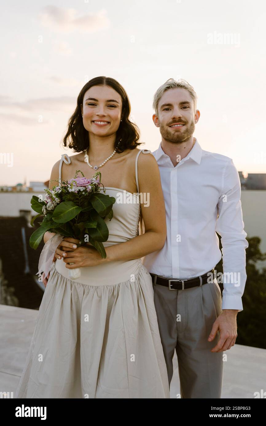 Portrait of smiling multiracial newlywed couple standing on rooftop ...