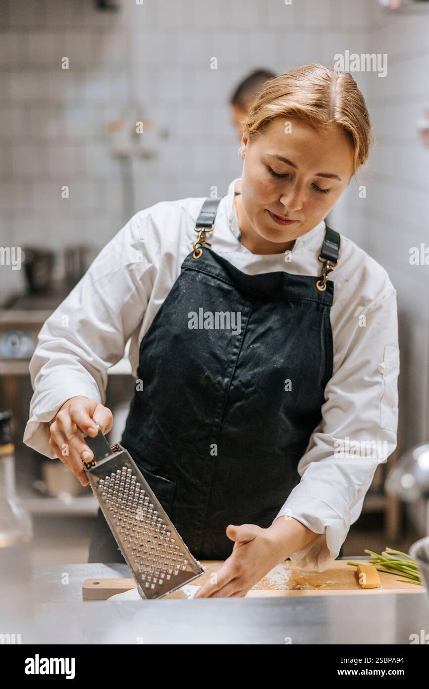 Front view of young female chef grating cheese with grater in kitchen ...