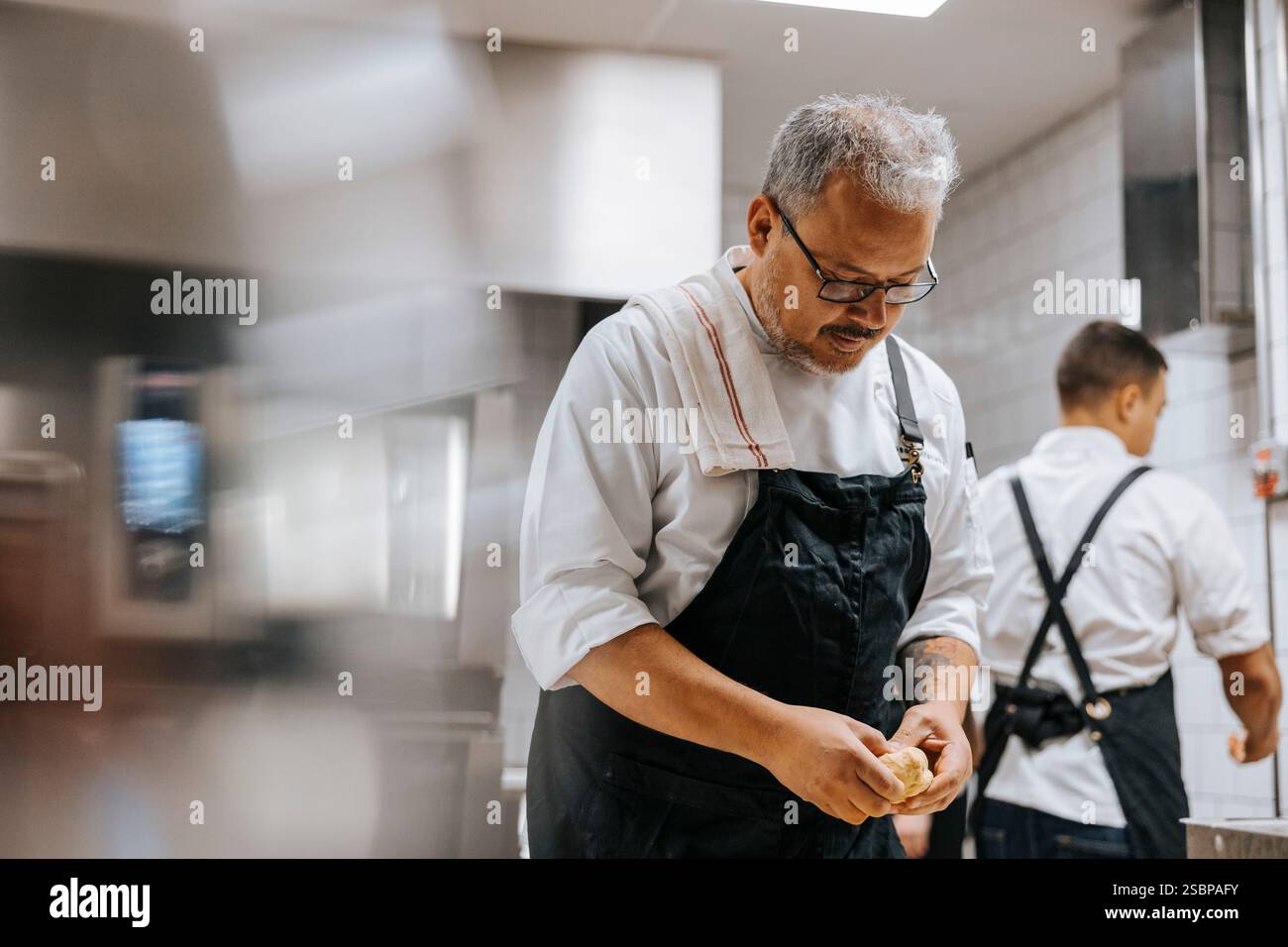 Mature male chef with eyeglasses kneading dough in restaurant kitchen ...