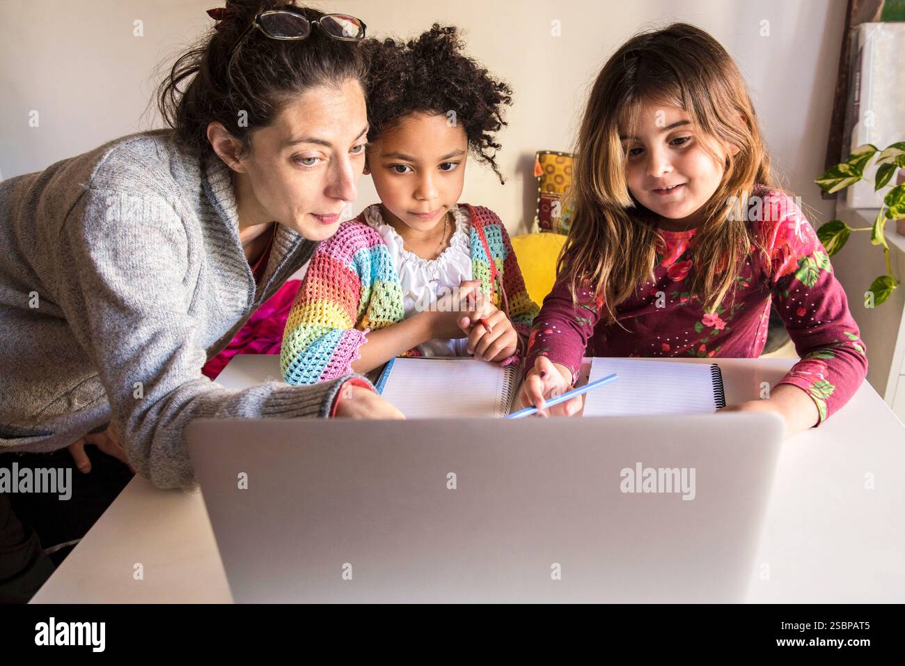 The woman is actively assisting two girls with their educational tasks at a table. They are ...