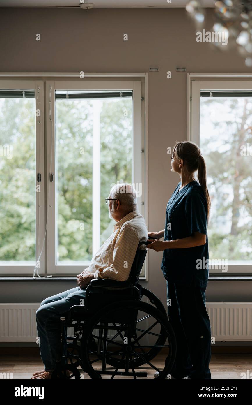 Female nurse with elderly male sitting in wheelchair looking out of window at nursing home Stock ...