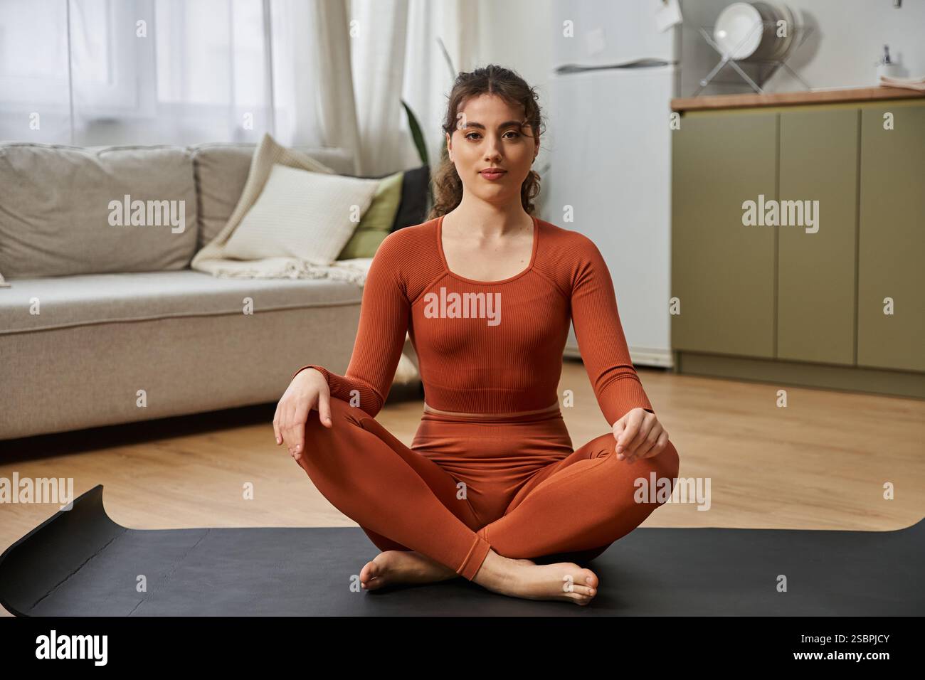 A beautiful young woman sits cross legged in a cozy living room, engaging in a calming yoga ...