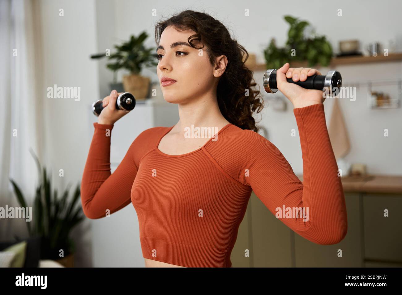 A beautiful young woman exercises with dumbbells in her cozy home ...