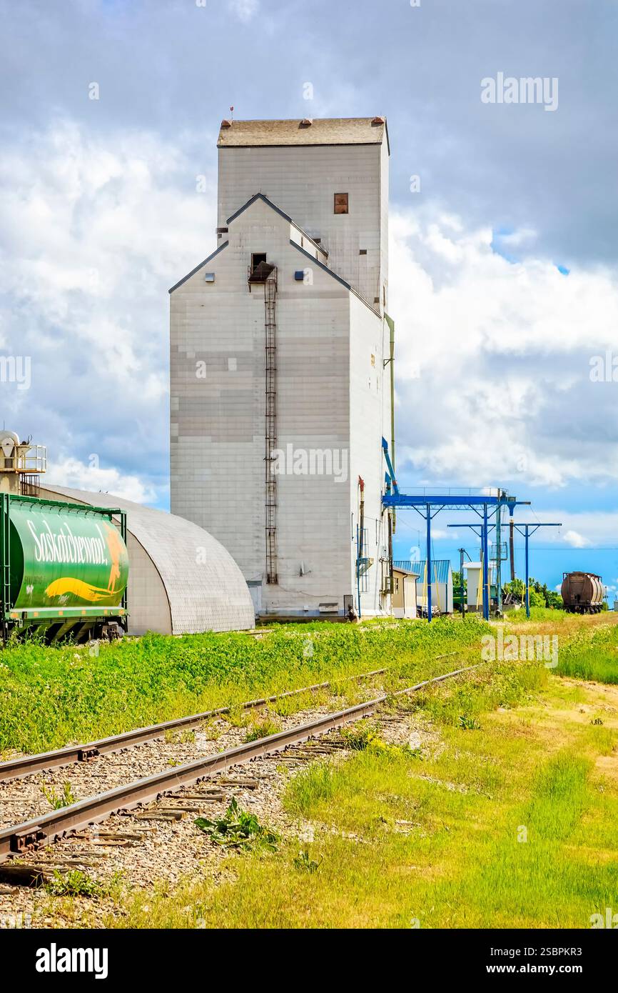 Train is on the tracks next to a grain silo. The train is green and has ...