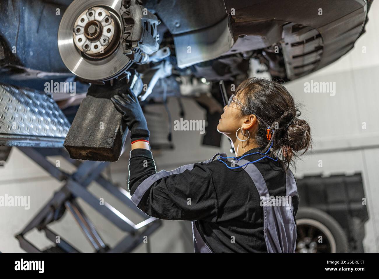 Skilled female mechanic working under a lifted car, inspecting and repairing the brake system in ...