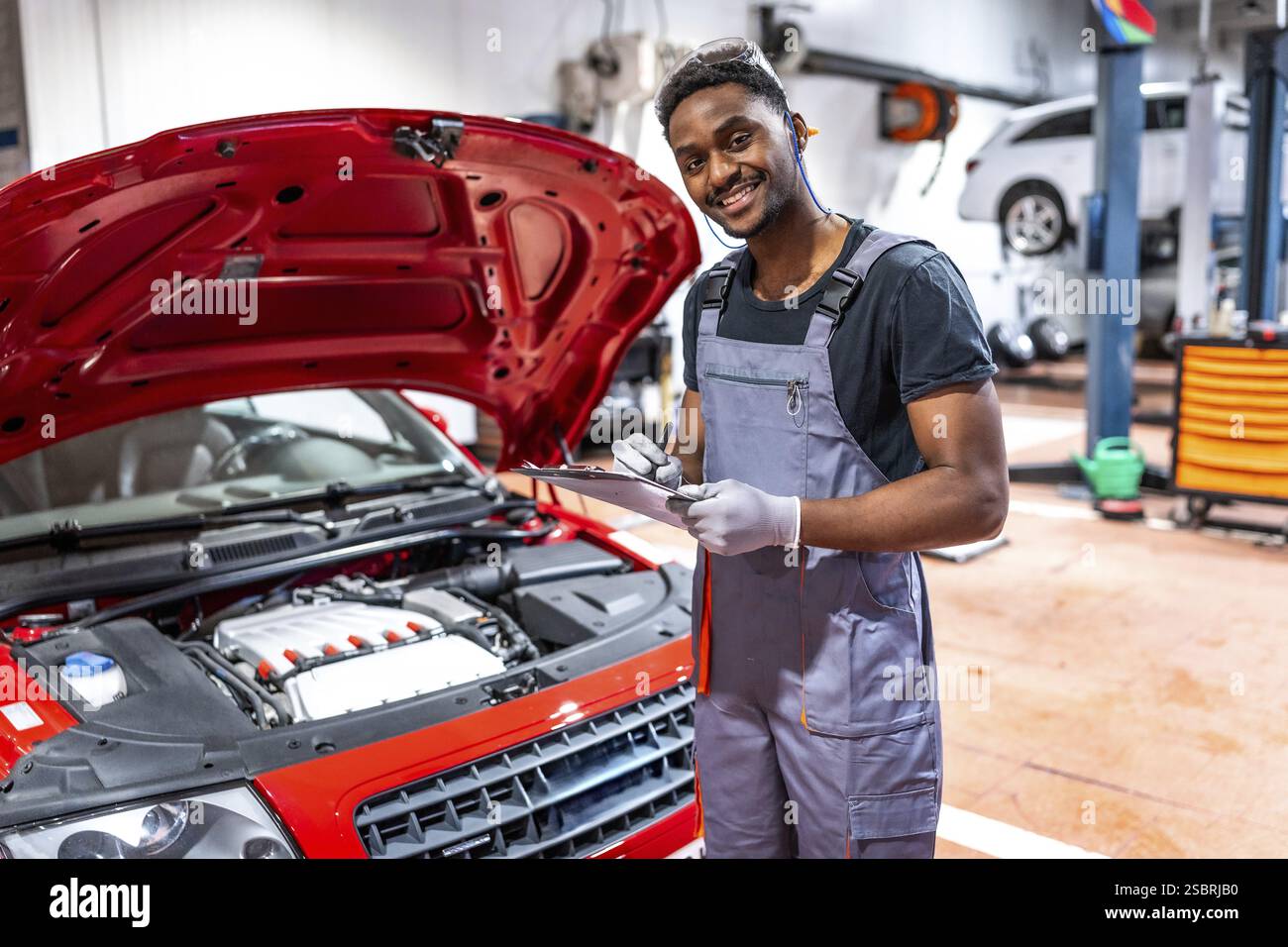 Smiling mechanic taking notes on clipboard while inspecting engine of ...