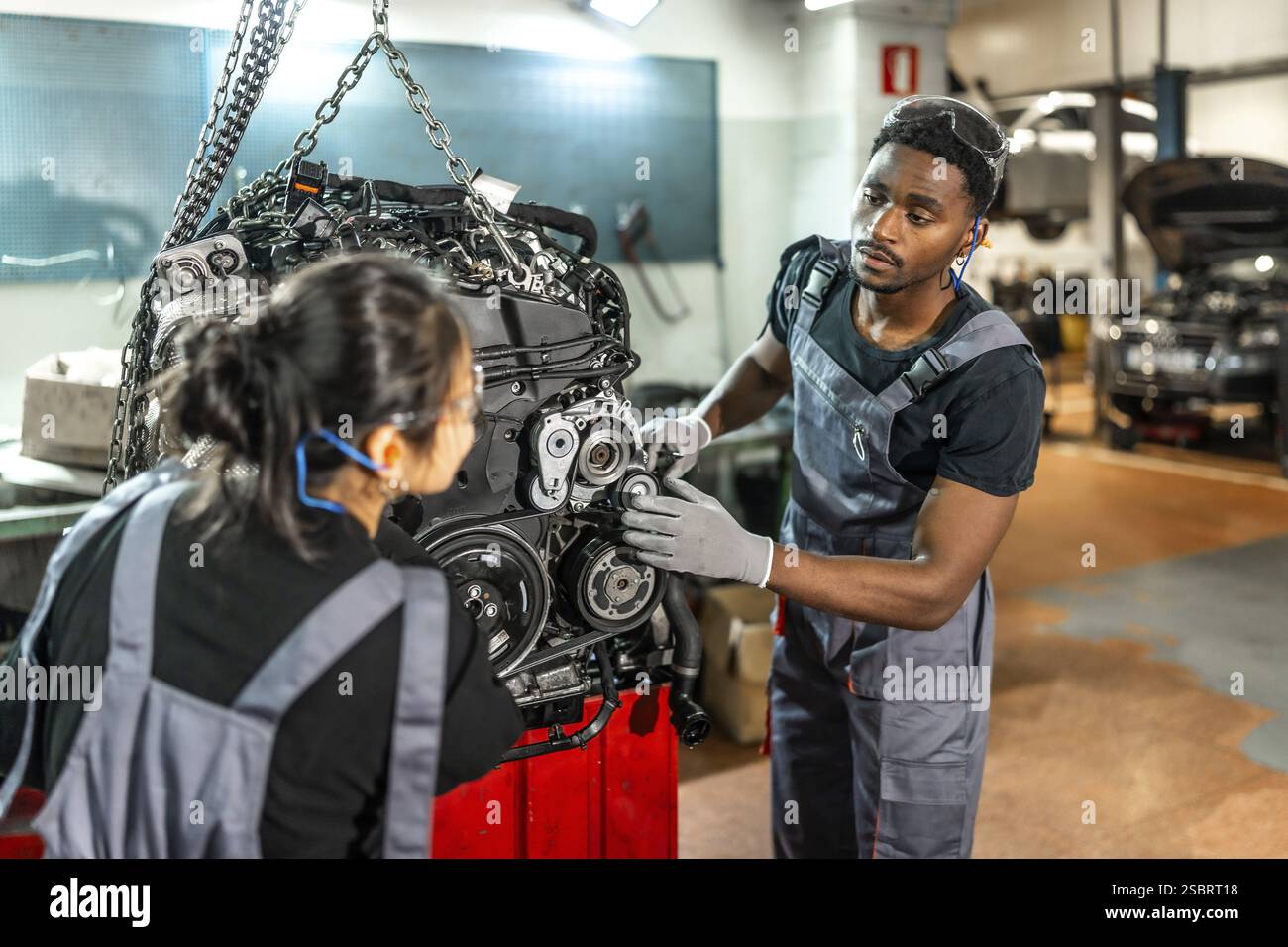 Two focused mechanics examining a car engine hanging from a crane in a ...