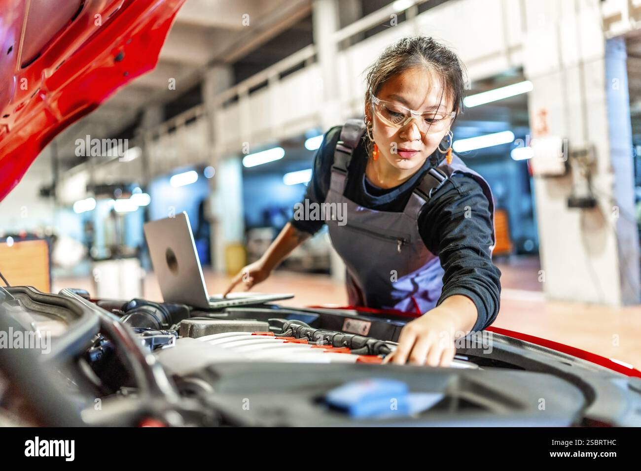 Young woman wearing mechanic overalls inspecting car engine using a laptop in a modern workshop ...