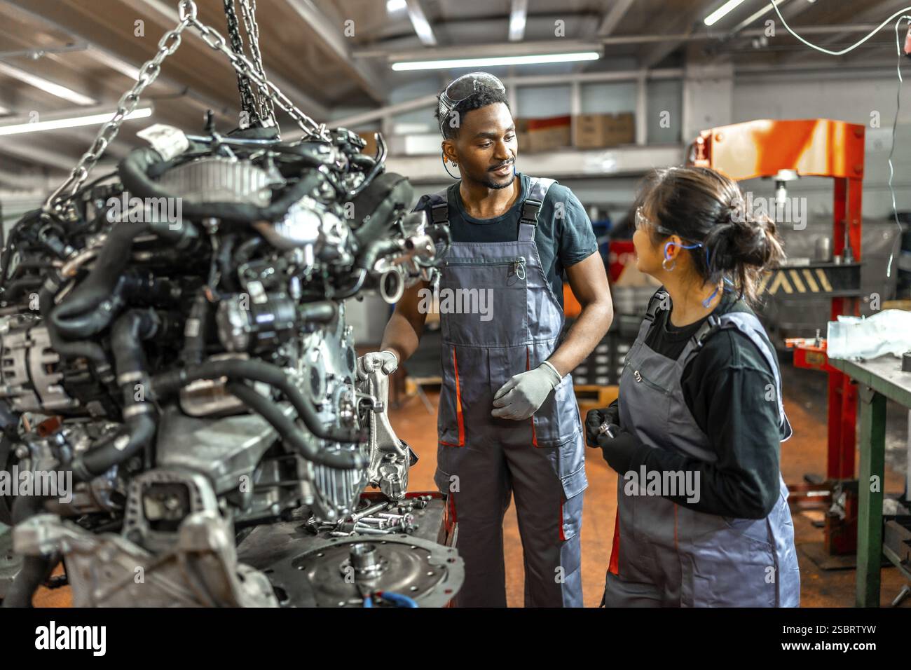 Two auto mechanics, a man and a woman, examining a car engine suspended ...