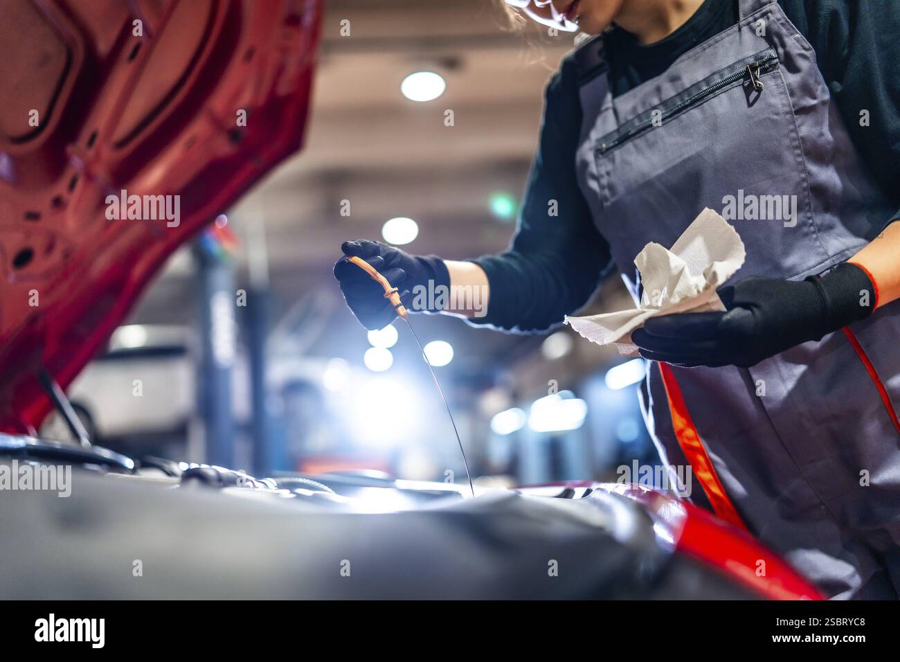 Professional female mechanic inspecting engine oil level with dipstick and rag in a car repair ...