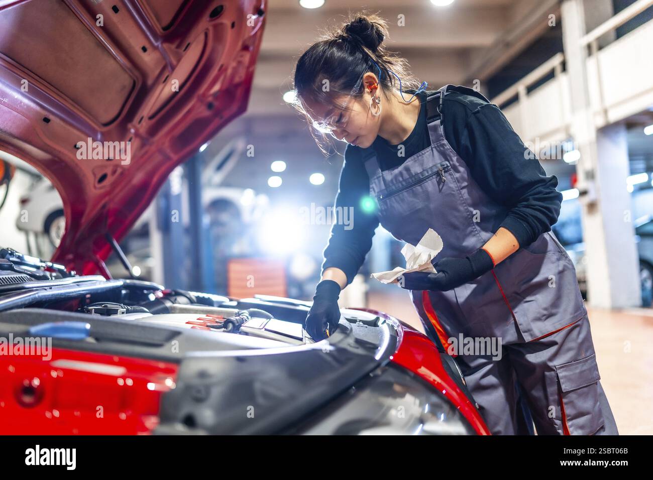 Young woman mechanic inspecting the engine oil level of a red car while working diligently in an ...